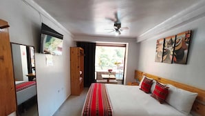 Quarto com cama, ventoinha no teto, TV, guarda-roupa de madeira, varanda com mesa e cadeiras, e obras de arte na parede.