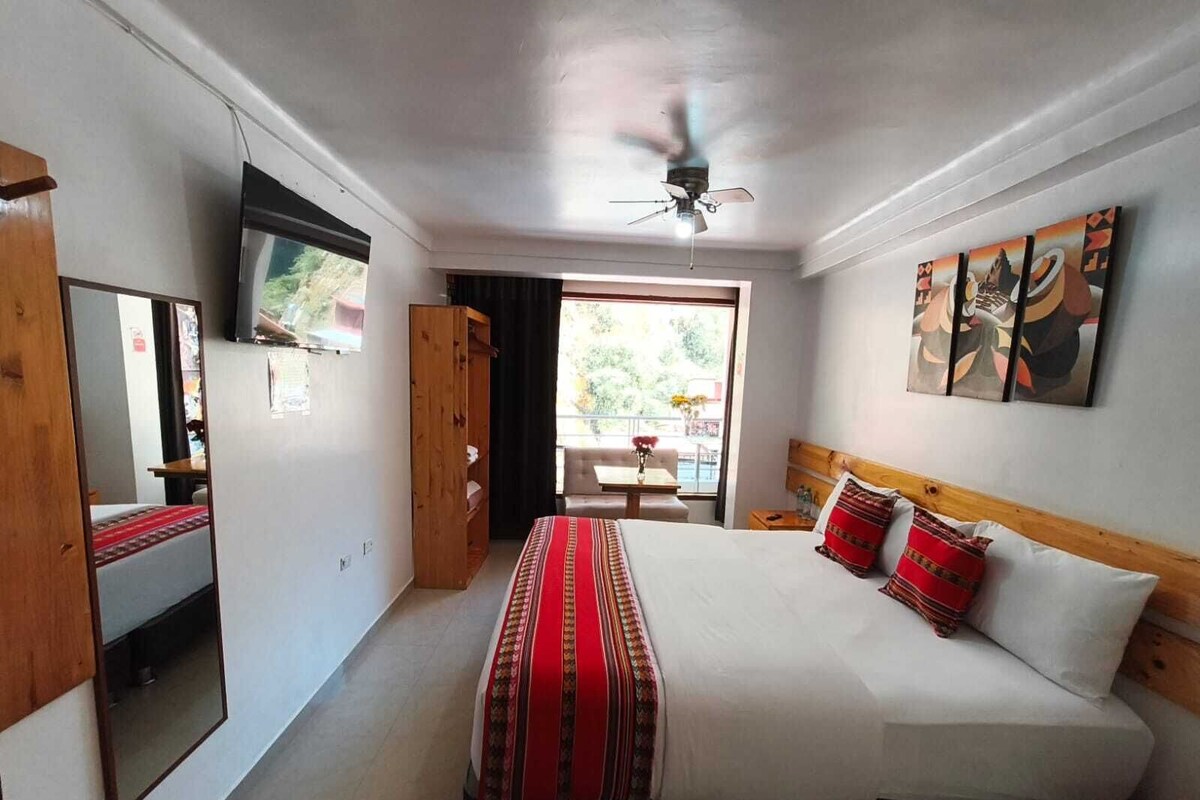 Quarto com cama, ventoinha no teto, TV, guarda-roupa de madeira, varanda com mesa e cadeiras, e obras de arte na parede.