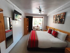 Quarto com cama, ventoinha no teto, TV, guarda-roupa de madeira, varanda com mesa e cadeiras, e obras de arte na parede.