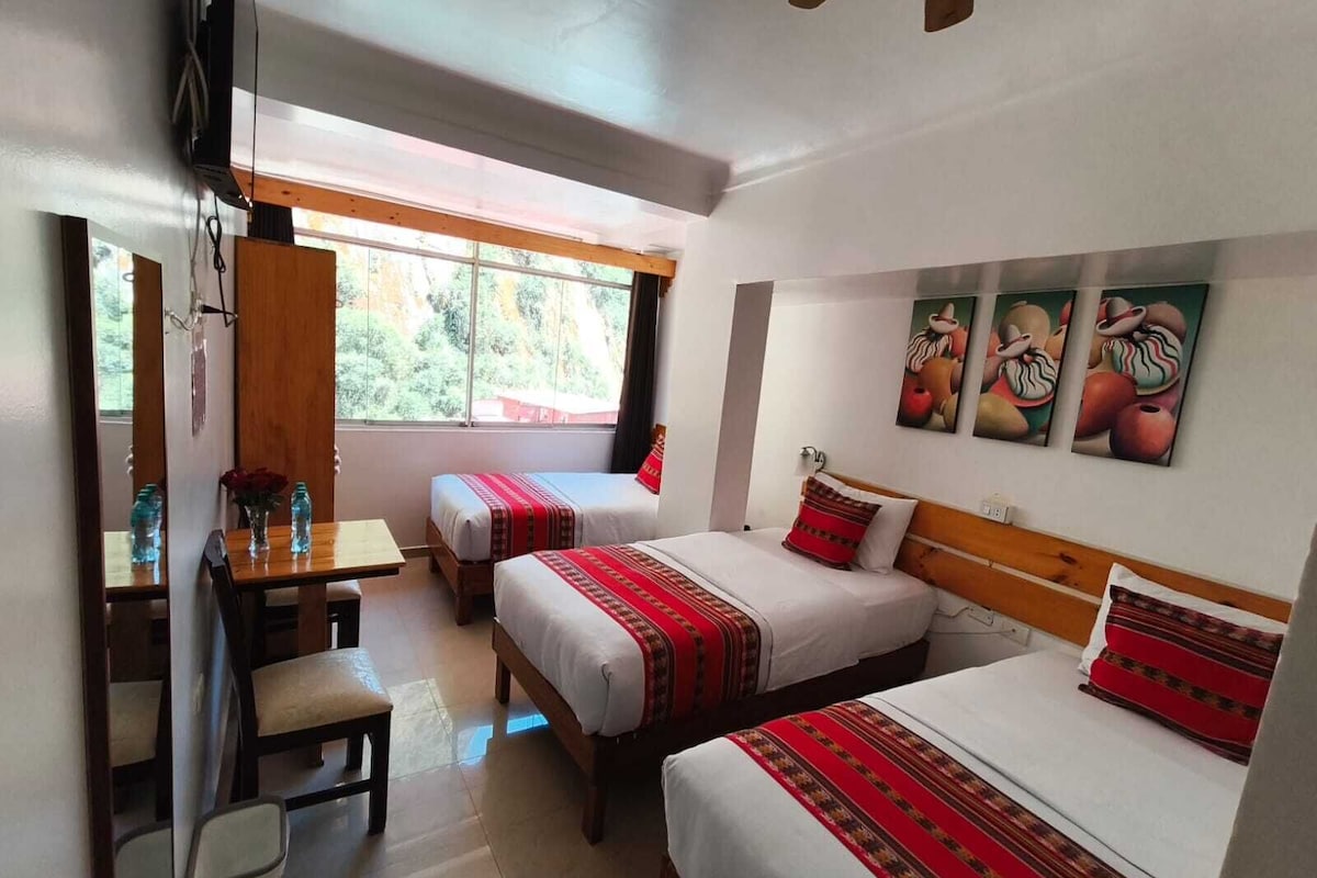 Quarto de hotel com duas camas, uma mesa e janela com vista para árvores.