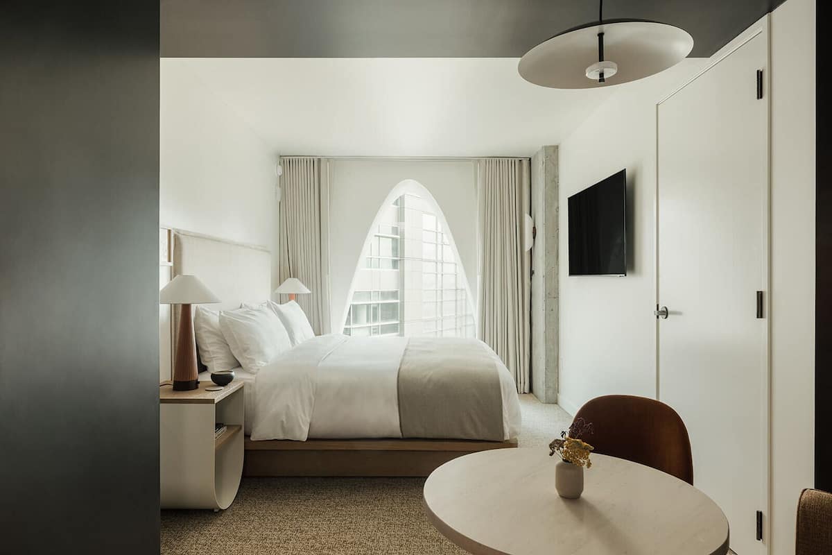 Quarto de hotel moderno com uma cama grande, uma mesa de cabeceira com luminárias, uma mesa redonda com um vaso e uma TV na parede.