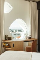 Quarto de hotel com escrivaninha curva, uma cadeira, uma cama, vista para a cidade e uma janela com cortinas.