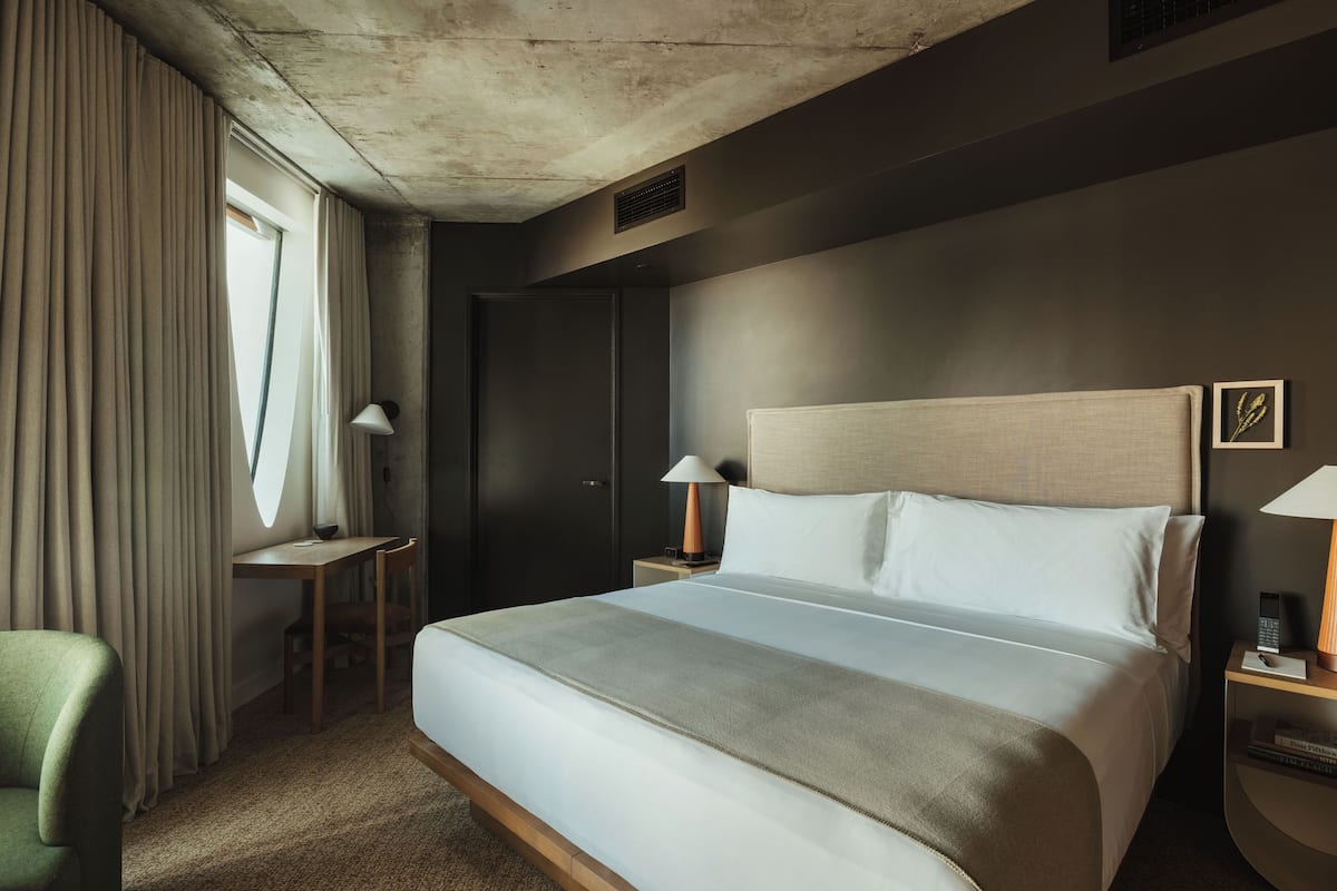 Quarto de hotel com uma cama grande, uma mesa, uma cadeira, um guarda-roupa e uma mesa de cabeceira com abajur.