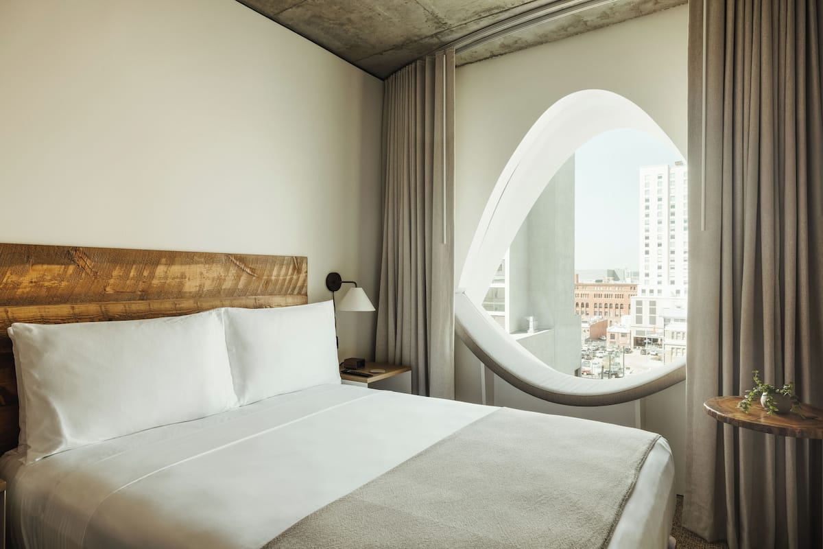 Quarto de hotel com uma cama grande, uma janela redonda com vista para a cidade, cabeceira de madeira e mesa de cabeceira com abajur.