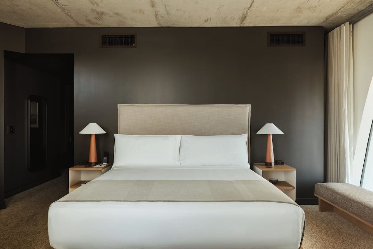 Quarto de hotel moderno com uma cama grande, duas mesas de cabeceira com luminárias, um banco e vista para o exterior.
