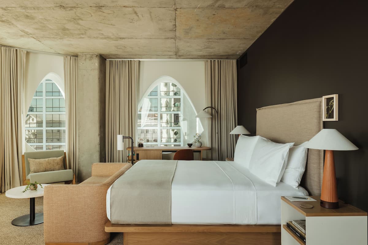 Quarto de hotel com uma cama grande, uma escrivaninha, uma cadeira e vista para um edifício através de janelas em arco.