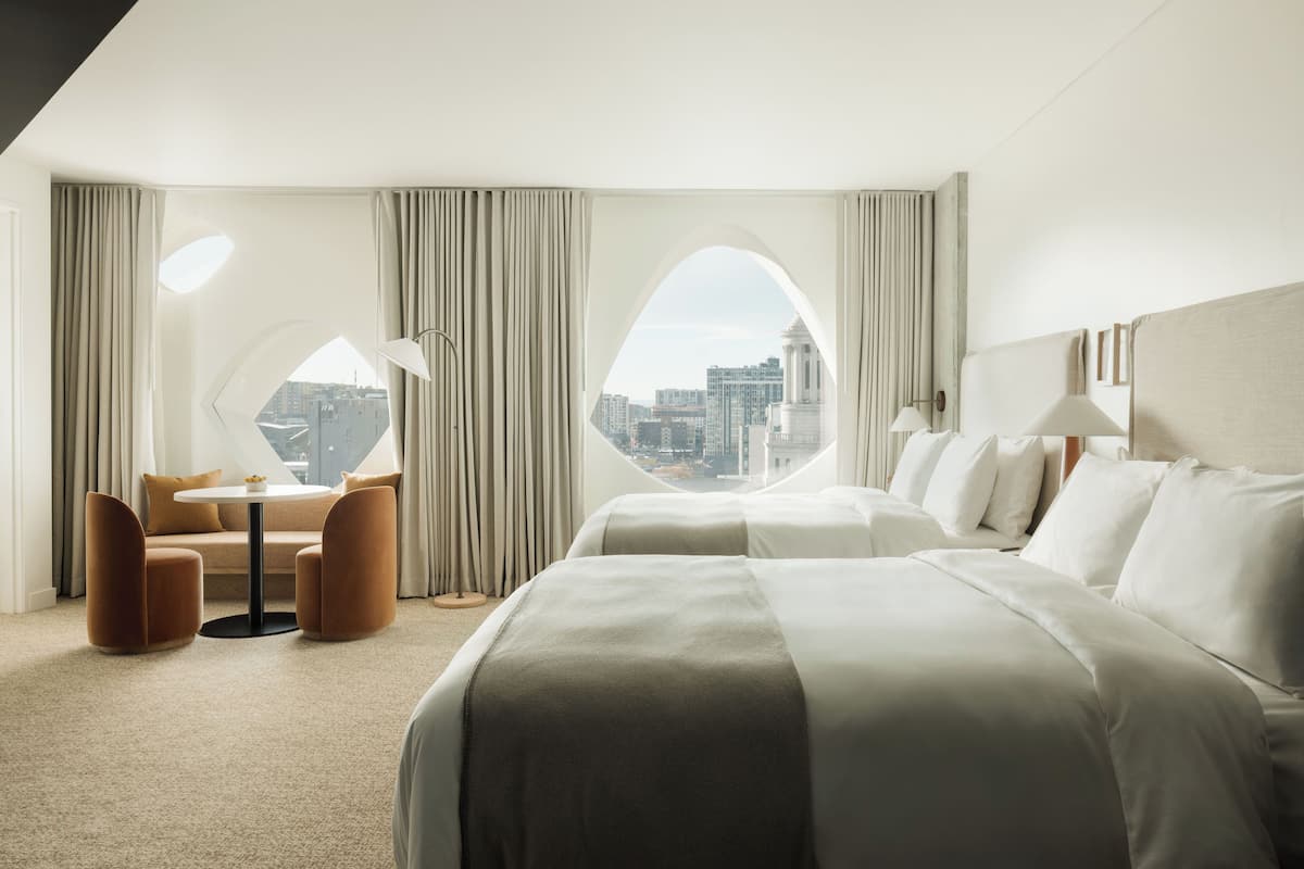 Quarto de hotel com duas camas, uma mesa redonda com cadeiras e uma janela grande com vista para a cidade.