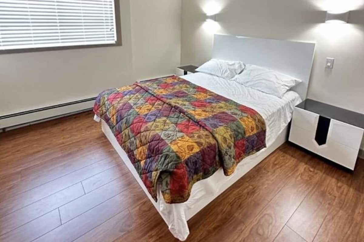 Quarto com piso de madeira, cama com colcha colorida, cabeceira branca e mesa de cabeceira com gaveta.