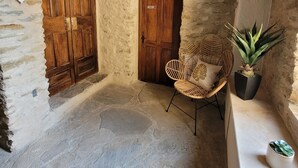 Um quarto rústico com paredes de pedra, duas portas de madeira, uma cadeira de vime com almofada e um vaso com planta.