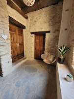 Um quarto rústico com paredes de pedra, duas portas de madeira, uma cadeira de vime com almofada e um vaso com planta.