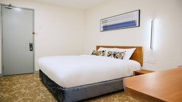 Quarto de hotel com cama, mesa de cabeceira de madeira, escrivaninha, tapete estampado, porta e quadro na parede.