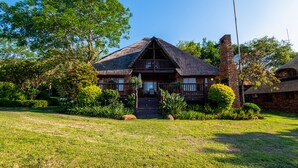 Uma casa com telhado de palha e varanda frontal, rodeada por vegetação exuberante e árvores.