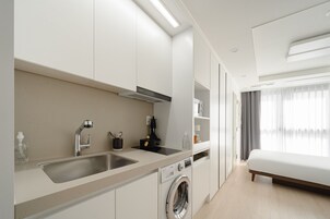 Quarto de hotel moderno com cozinha compacta, máquina de lavar, cama e uma grande janela com cortinas.