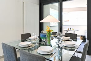 Uma zona de refeições moderna, com mesa de vidro, cadeiras cinzentas e um vaso com flores.