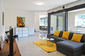 Uma sala de estar moderna com um sofá cinzento, almofadas amarelas, uma pintura e um espaço de jantar com mesa e cadeiras.