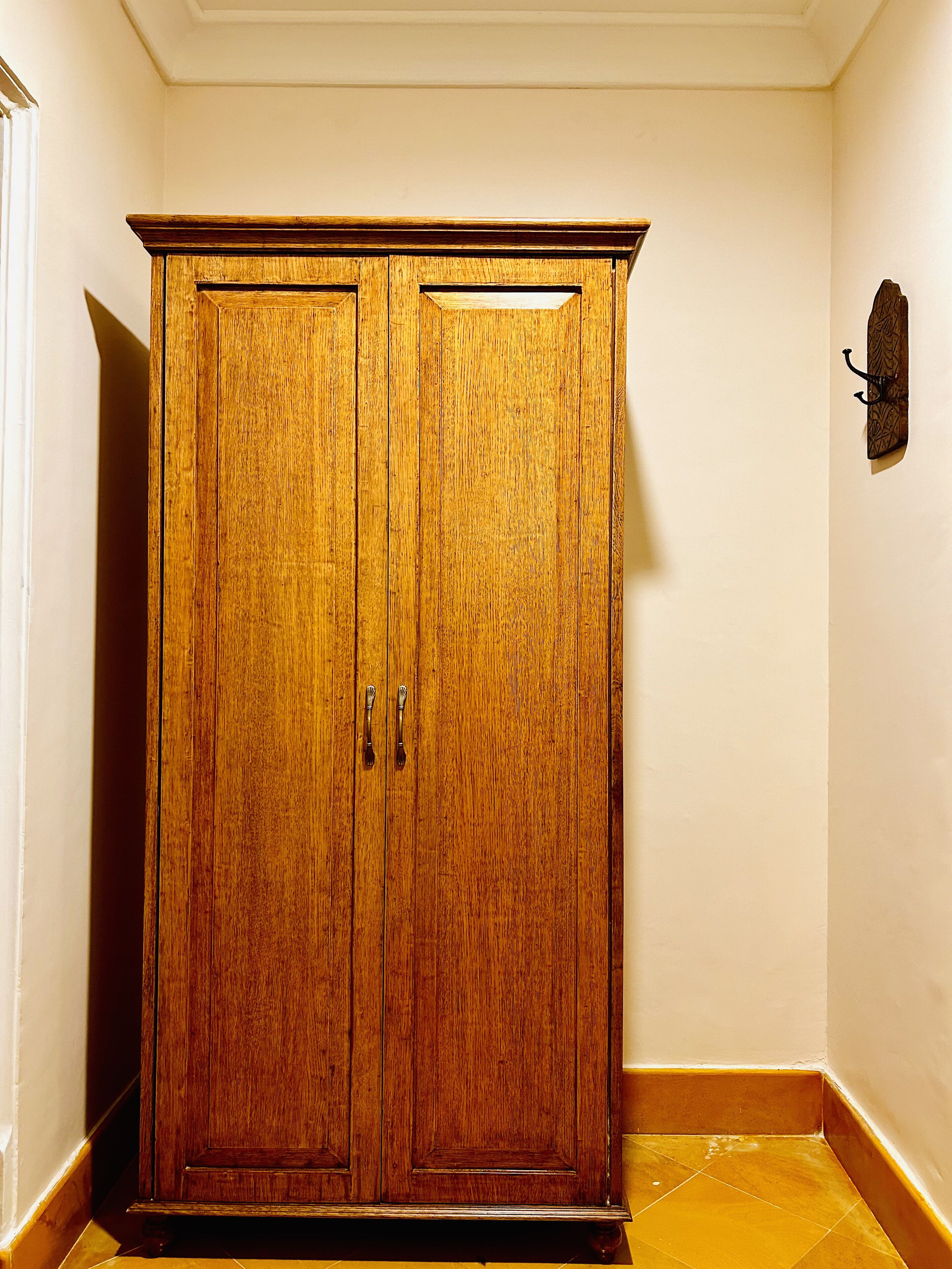 Guarda-roupa de madeira com duas portas e uma pequena prateleira, localizado num canto de um quarto com paredes simples e chão em azulejos.