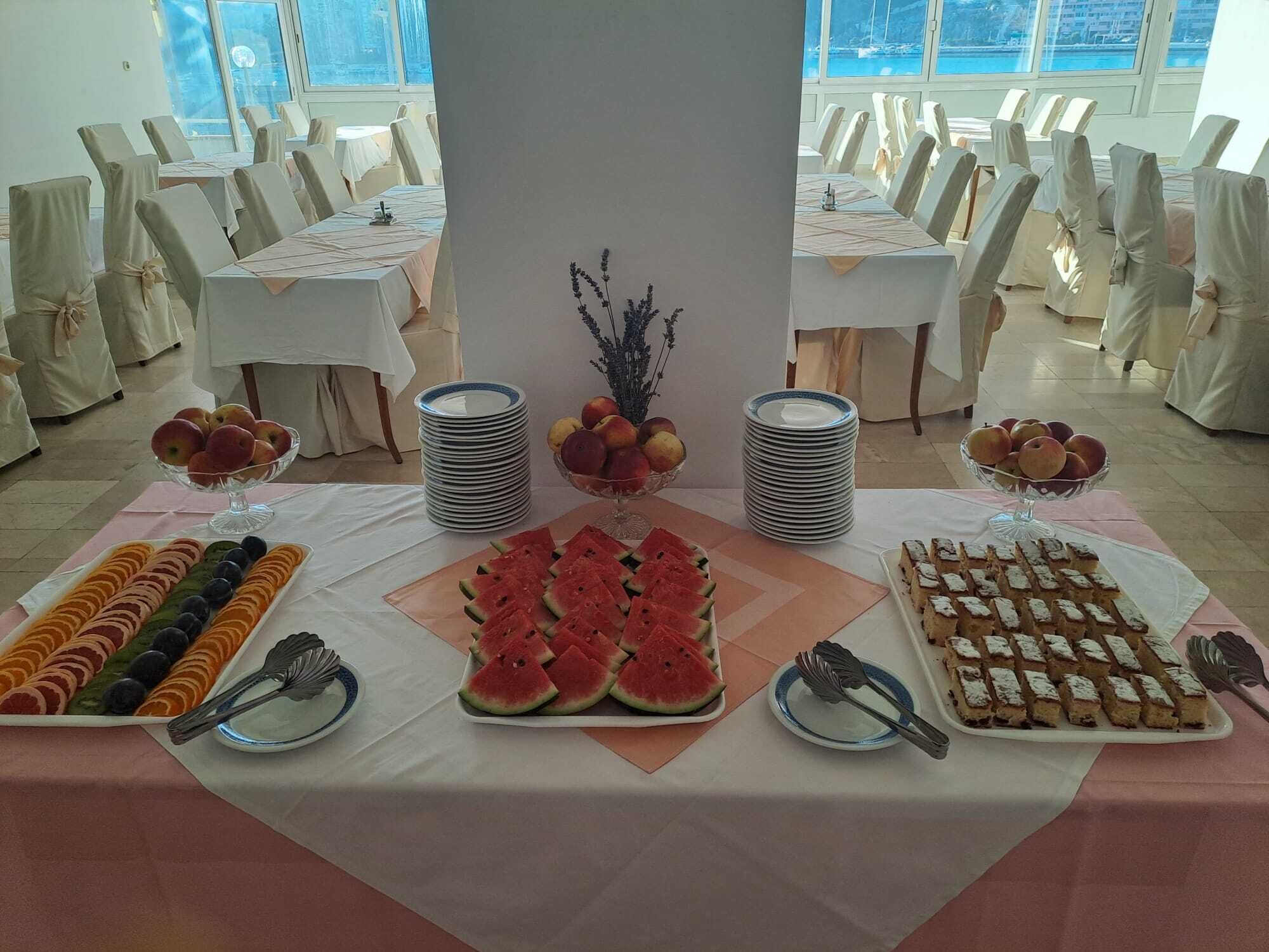 Mesa de buffet com diversas travessas de fruta, incluindo melancia e pêssegos, e uma seleção de sobremesas.