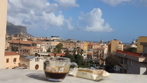 Paisagem urbana com edifícios, céu claro e uma xícara de café sobre uma mesa.