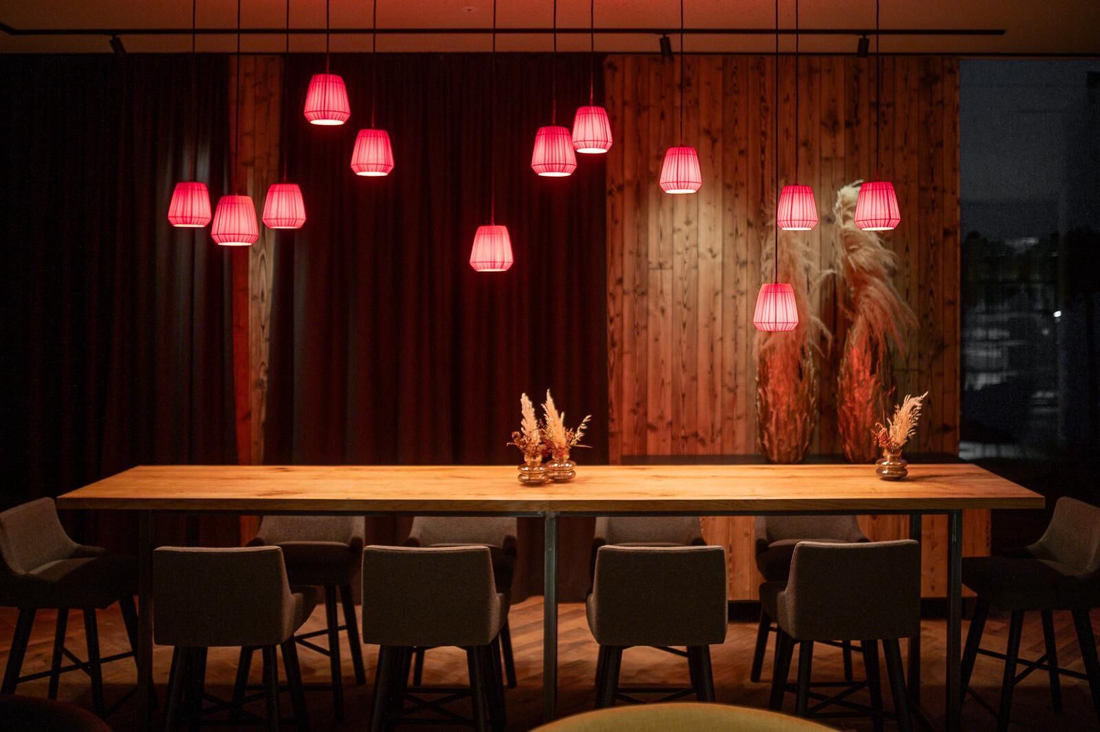Uma sala de jantar com uma longa mesa de madeira, seis cadeiras e várias luminárias vermelhas penduradas.
