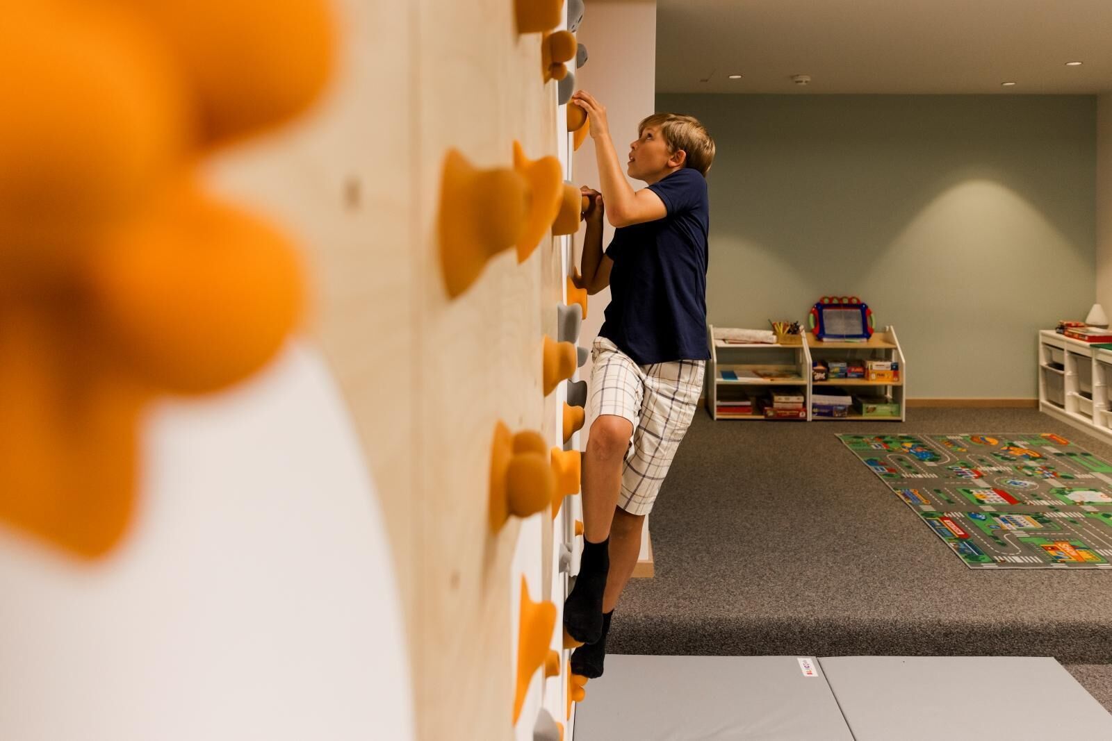 Uma criança escalando uma parede com apoios laranja em um quarto com piso coberto por carpete, prateleiras com livros e um tapete de brincadeira.