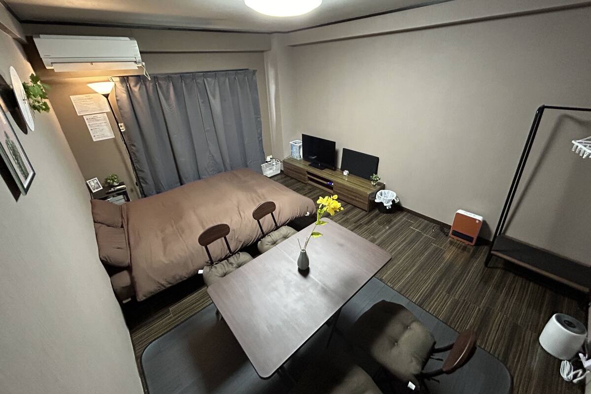 Quarto de hotel pequeno com uma cama, uma mesinha com um vaso, uma cadeira, uma TV e uma escrivaninha.