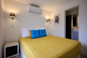 Quarto de hotel com cama, duas luminárias fixadas na parede, ar condicionado e um quadro emoldurado.