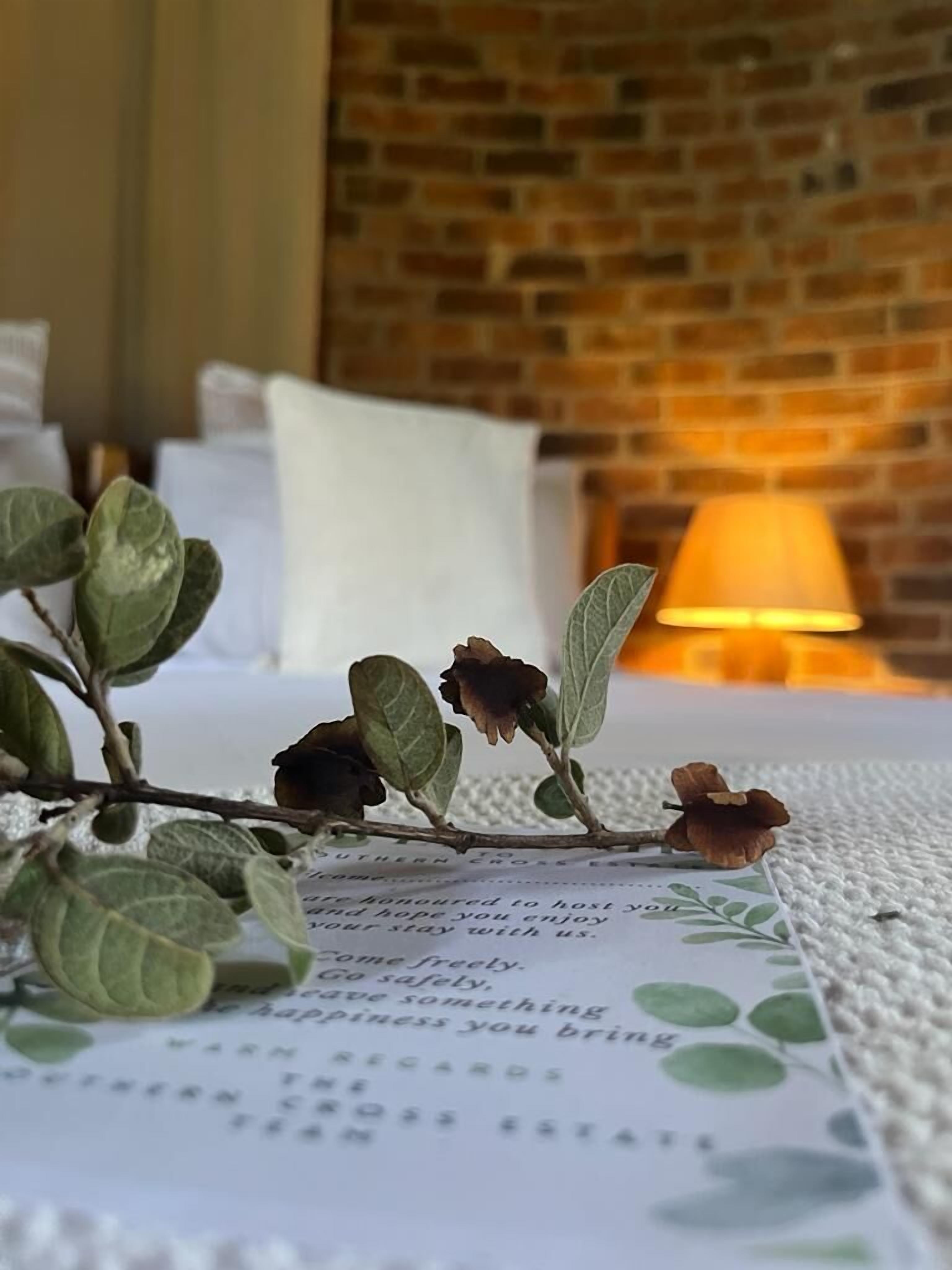 Um ramo com flores secas sobre uma mesa com uma mensagem impressa, ao fundo uma parede de tijolos e um candeeiro aceso.