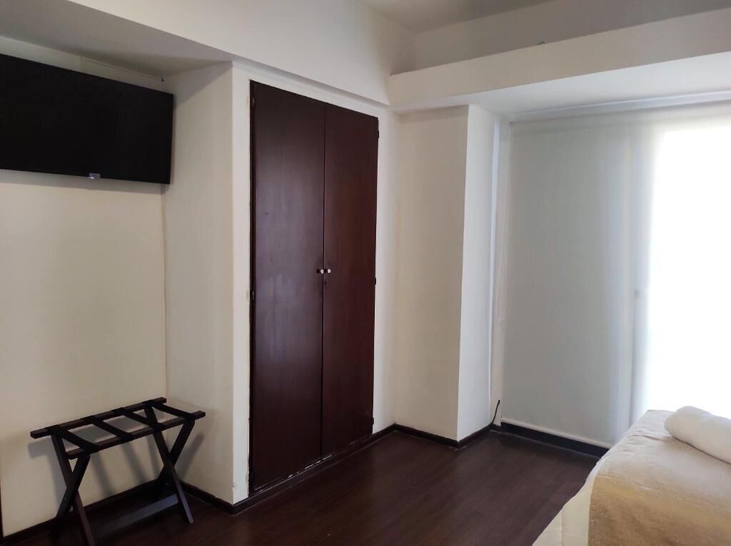 Quarto com armário de madeira escura, uma pequena mesa e uma cama com almofada branca.