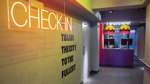 Um hall de hotel com um letreiro de neon que diz "CHECK-IN TO LIVE THE FULLEST" e um balcão de recepção com dois alto-falantes e um monitor de computador.