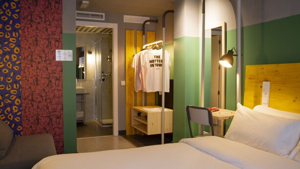 Quarto de hotel com cama, escrivaninha, cadeira, espelho e banheiro visível através de uma porta aberta.