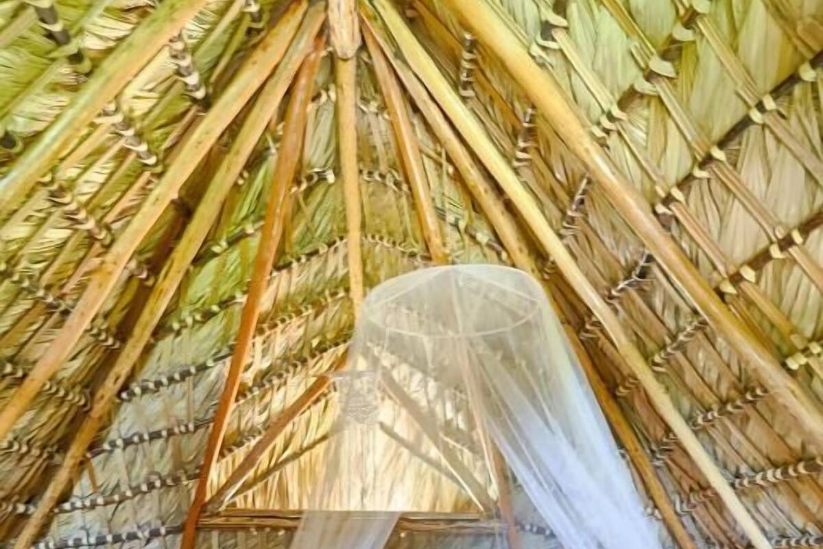 Tenda com telhado de palha, uma cama, uma mesa pequena e um objeto decorativo.