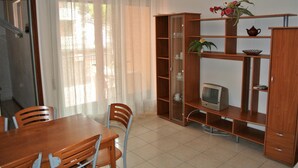 Uma área de refeições com uma mesa e cadeiras de madeira, uma televisão sobre uma prateleira e uma janela com cortinas leves.
