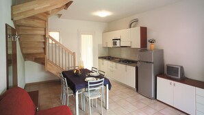 Cozinha com mesa de jantar, cadeiras, geladeira, micro-ondas e televisão. Escada de madeira leva a outro cômodo.