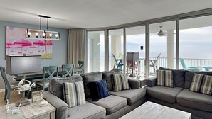 Uma sala de estar moderna com um sofá cinza, uma mesa de centro e um espaço de jantar com cadeiras e uma mesa. Grandes janelas oferecem vista para o oceano.