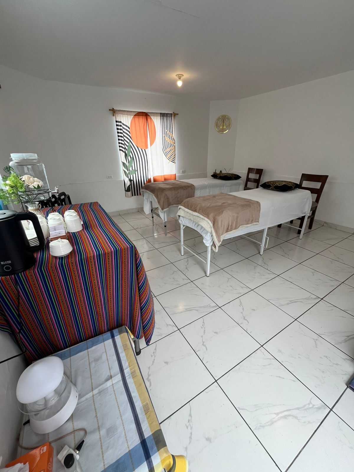 Quarto com duas camas, uma mesa pequena com toalha de mesa listrada e colorida, uma máquina de café e uma janela com cortina.
