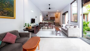 Uma sala de estar ampla com sofá, mesa de centro e área de jantar. Um quadro está na parede, e cozinha ao fundo.