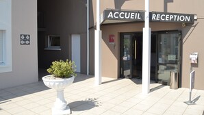Uma área de recepção com uma planta em vaso ao primeiro plano e uma placa sobre a entrada com a inscrição "ACCEIL RECEPTION".