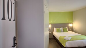 Quarto de hotel com cama, almofadas verdes, colcha branca e uma parede verde de destaque.
