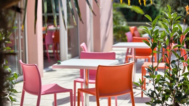 Uma área de pátio externa com cadeiras e mesas em tons de rosa e laranja, cercada por vegetação.