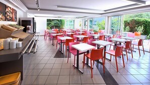 Uma sala de jantar moderna com mesas e cadeiras vermelhas e brancas, janelas amplas e um balcão com alimentos.