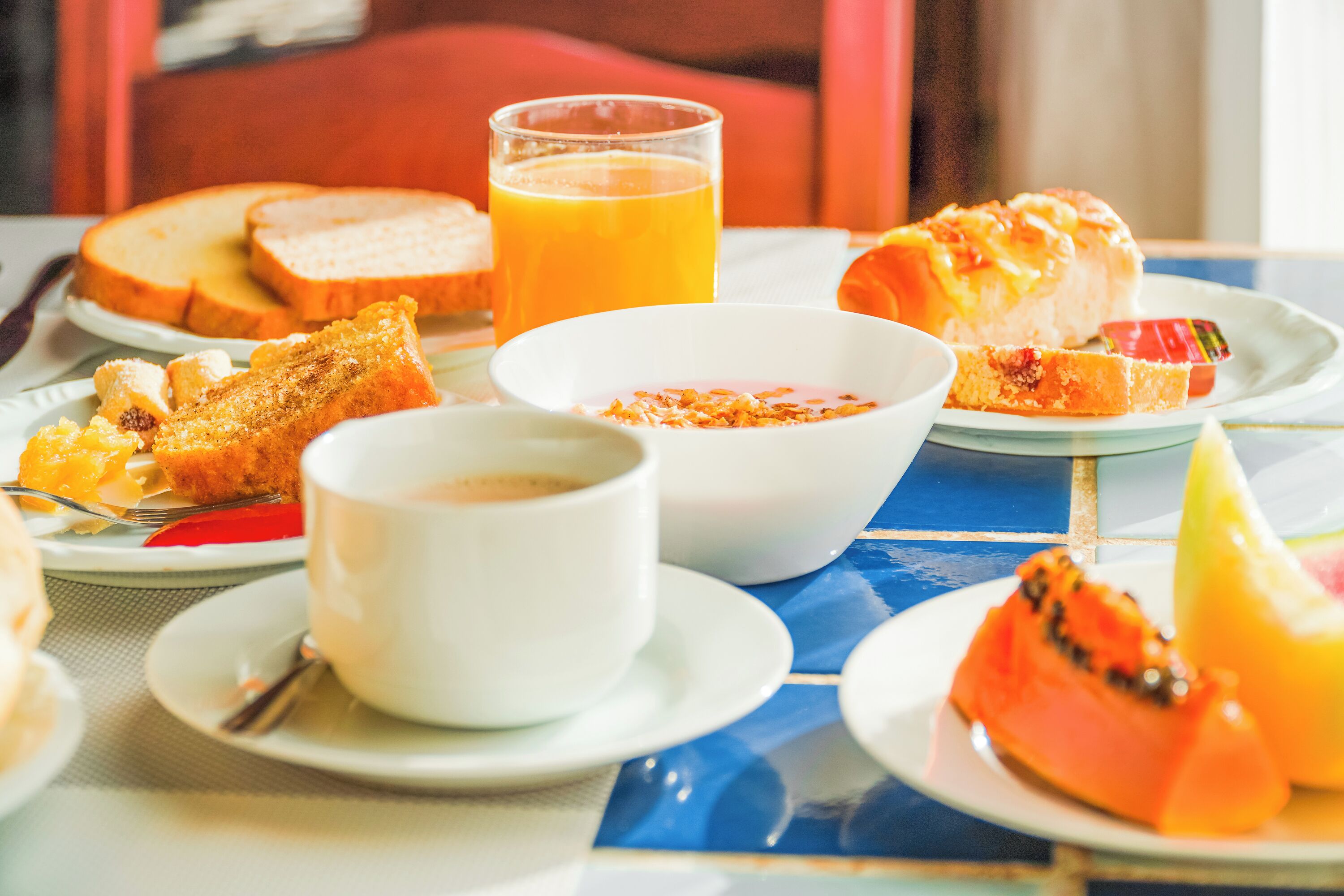 Uma mesa de café da manhã com torradas, um prato de cereal, um copo de suco de laranja, uma fatia de bolo e uma mamão.