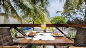 Uma mesa de madeira com um jogo americano branco, duas cadeiras de vime e um copo de bebida em uma varanda com vista para palmeiras e uma praia.