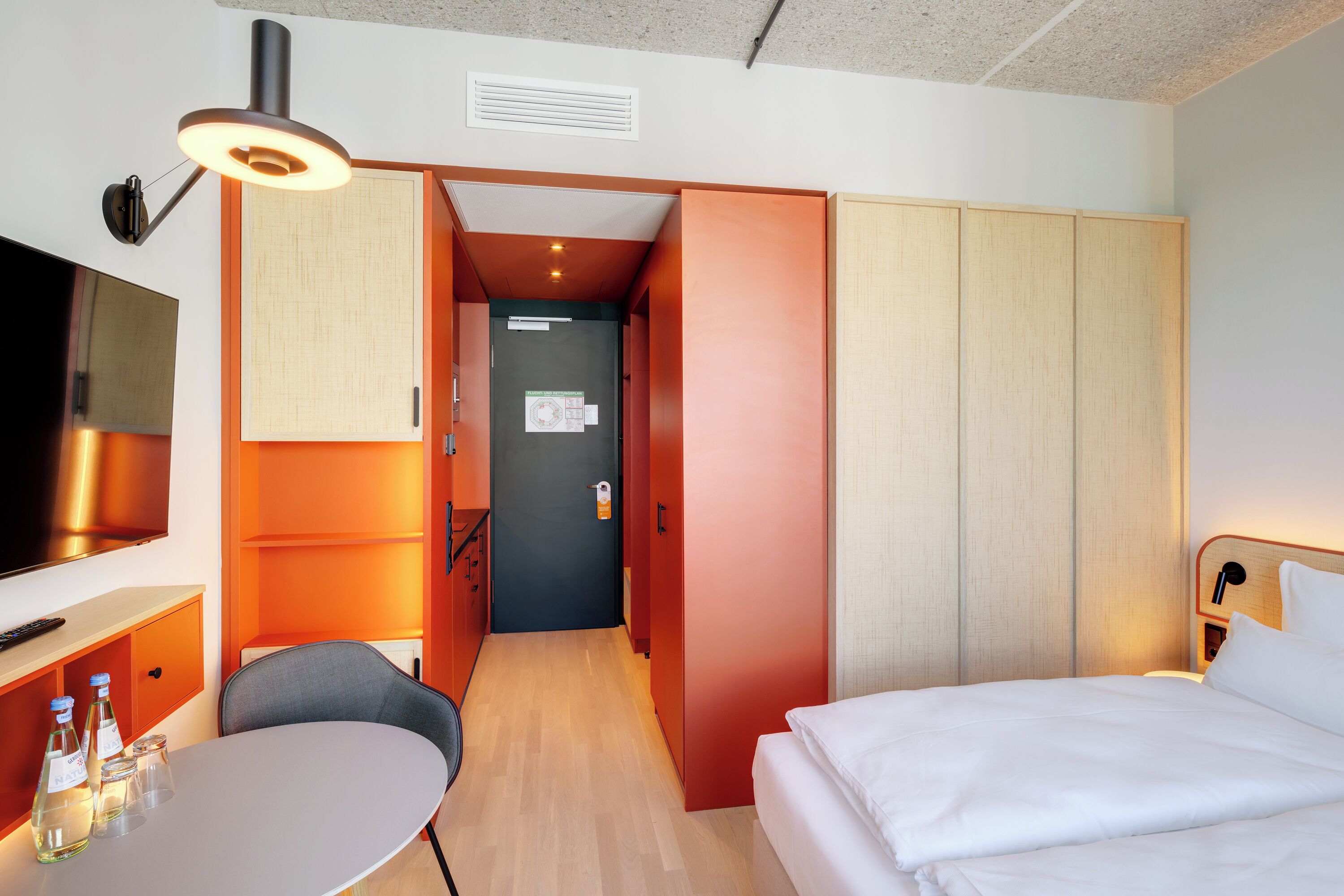 Quarto de hotel com cama, escrivaninha com cadeira, televisão e guarda-roupa.