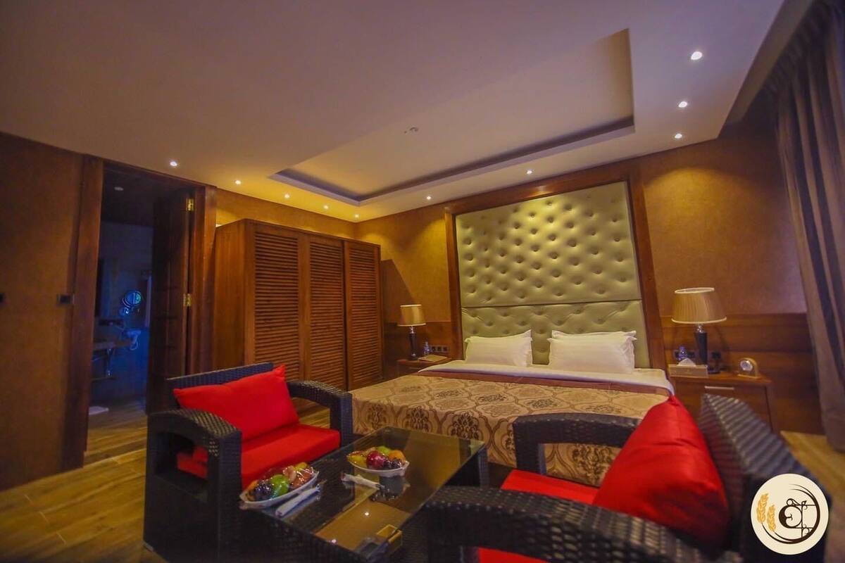 Quarto de hotel com uma cama grande, duas poltronas, uma mesa de centro com frutas e um guarda-roupa.