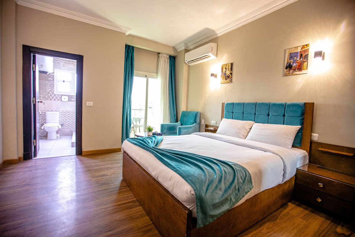 Quarto com cama de madeira, mesa de cabeceira, cadeira e vista para o banheiro por uma porta aberta.