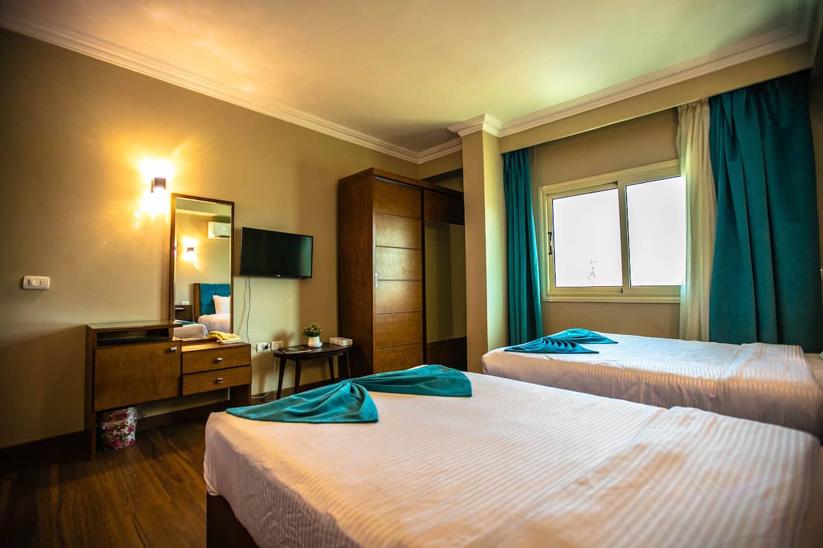 Quarto de hotel com duas camas, guarda-roupa de madeira, escrivaninha com cadeira, mesa pequena com planta e televisão na parede.