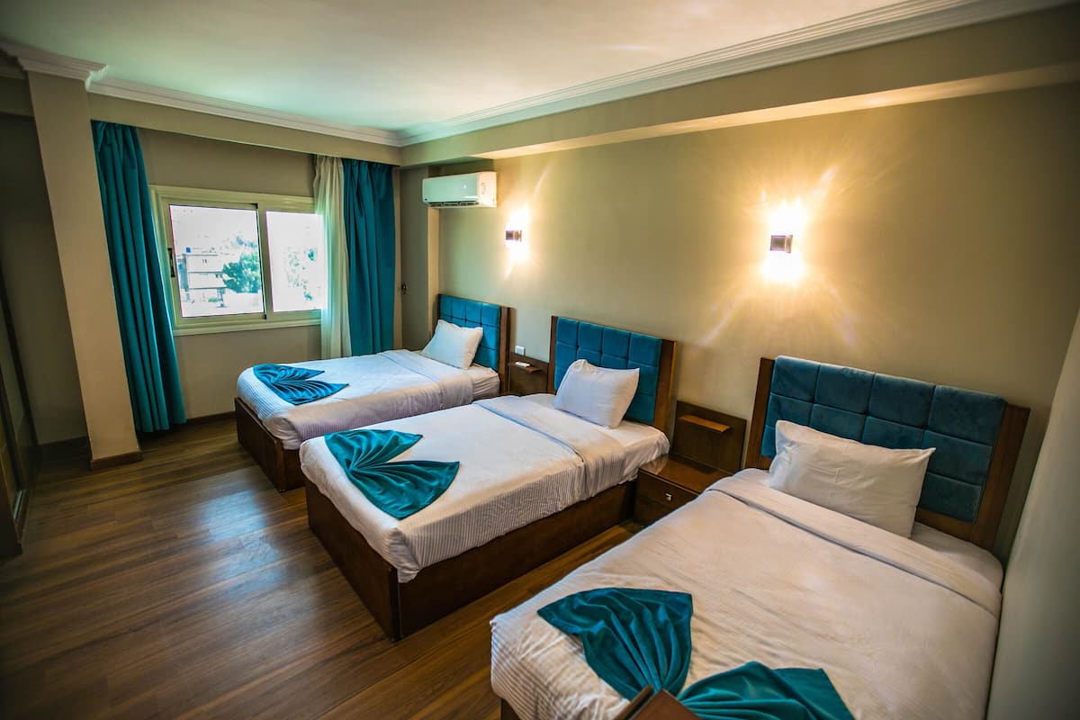Quarto de hotel com três camas, piso de madeira, cortinas verde-água e janela com vista.