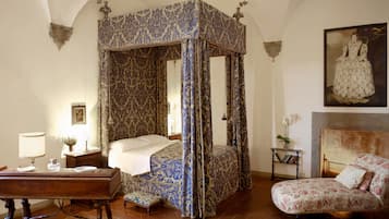 Uma cama de dossel com cortina florida, uma mesinha de cabeceira de madeira com abajur, uma cadeira com almofada florida e um quadro na parede.