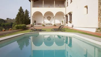 Uma área de piscina com espreguiçadeiras e uma mesa de jantar, cercada por um edifício com arcos e vista para as colinas.