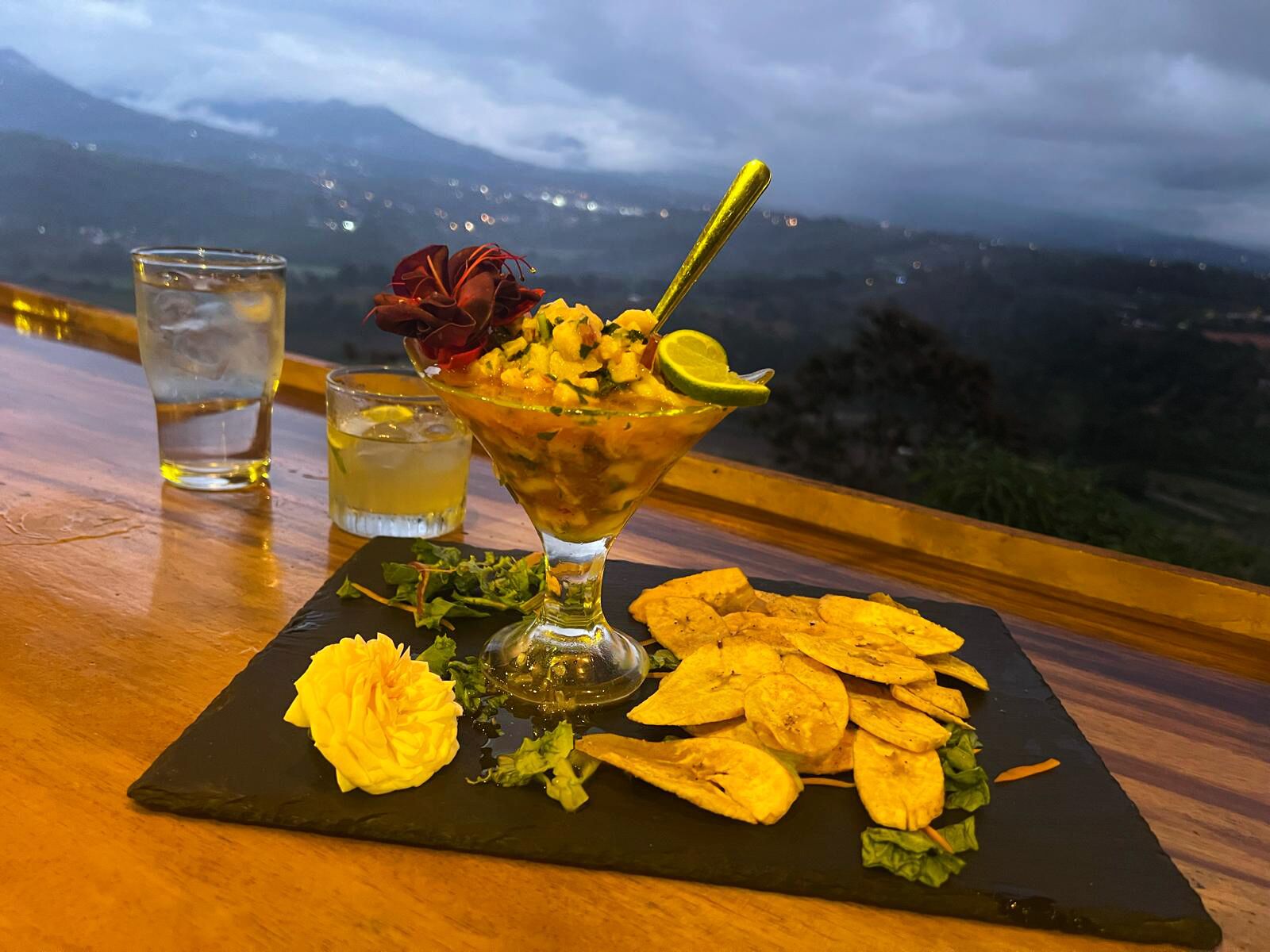 Uma mesa com uma bebida tropical, salgadinhos e uma flor sobre um ladrilho negro.